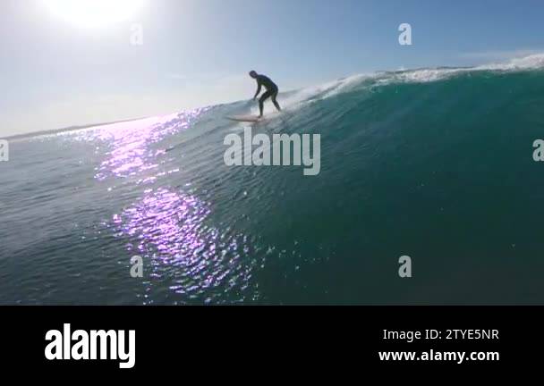 SLOW MOTION, LENS FLARE: Surfboarder riding a small wave on a bright ...