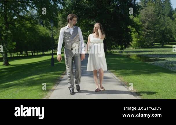 SLOW MOTION: Happy pregnant couple holding hands and walking trough green park in sunny spring ...