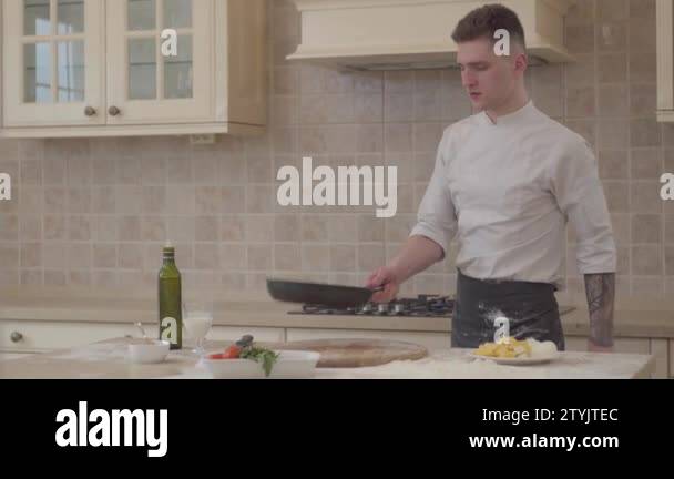 Young man in chef uniform puts salt in the fryng pan and throws up meat ...