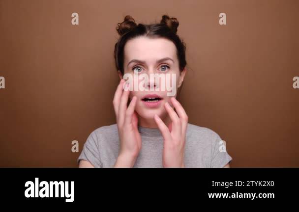Bewildered young brunette woman touches cheek, opens mouth, has eyes ...