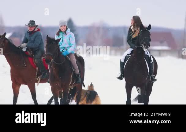 Three women riding horses in a village with a dog running near by them ...