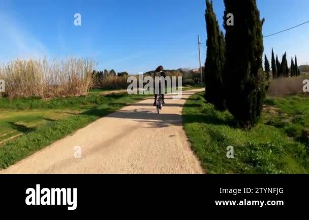 A girl with long hair rides a bicycle on a rural road among fields ...