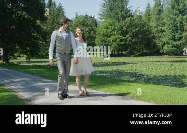 SLOW MOTION: Happy pregnant couple holding hands and walking trough green park in sunny spring ...