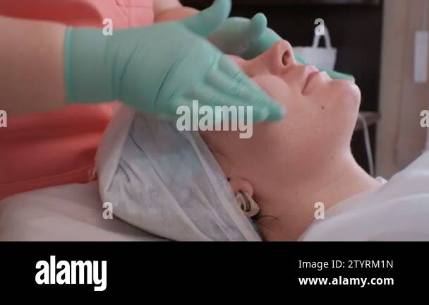 Beautician in blue gloves puts the face of a young woman lather ...