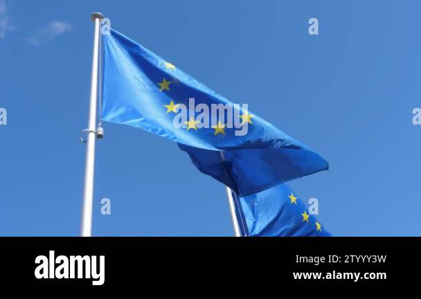 Two EU flags waving freely on flagpole in celeste sky in spring in slow ...