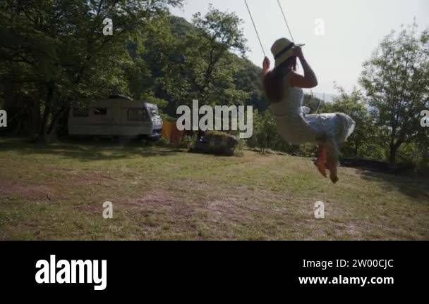 Young lady in romantic dress, hat swings on caravan camping. Side view ...