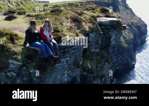 Two girls sit on the edge of the famous Cliffs of Moher in Ireland ...
