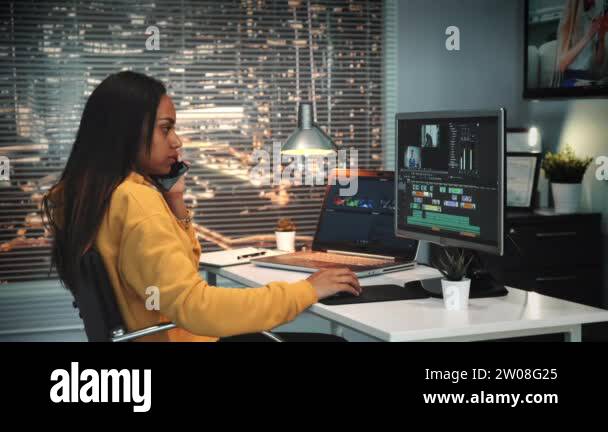 Back view of female video editor talking with boss by smartphone and working with footage and ...