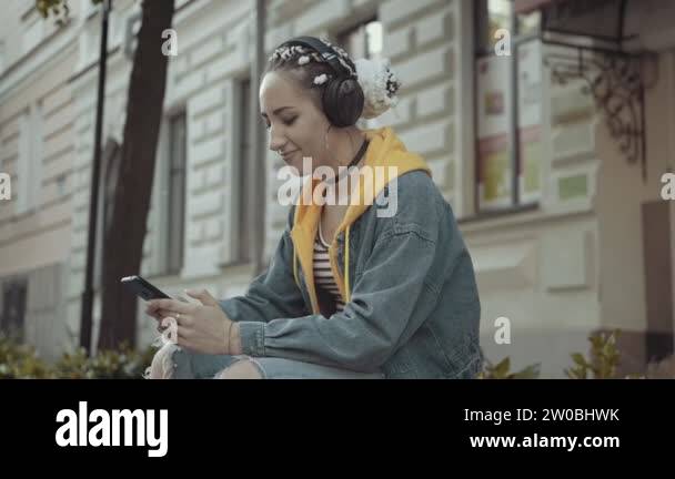 Stylish young woman texting and listening to music on phone outdoors ...