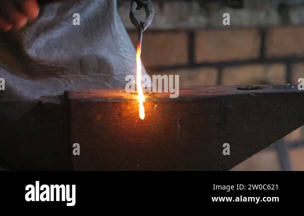 Blacksmith moving an iron rod in tongues over a steel anvil in a dim ...
