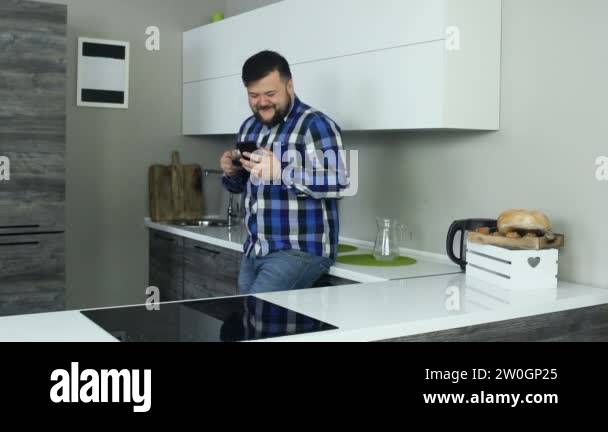 A stout man stands in the kitchen and laughs, looking at the phone ...