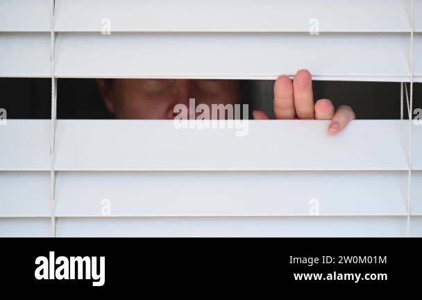 Suspicious person looking outside home window from behind Venetian ...