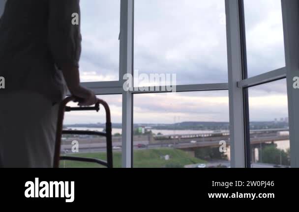 Weak old lady using walking frame, looking through nursing home window ...