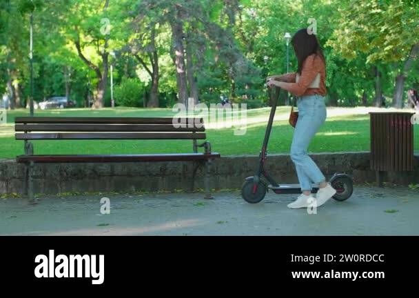 A young woman arriving in park on electric push scooter, sits on a park bench and reading book ...
