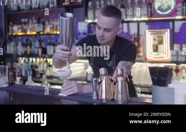 Young barman grating small kiwi on a grater over a glass of creamy cocktail standing on the bar ...