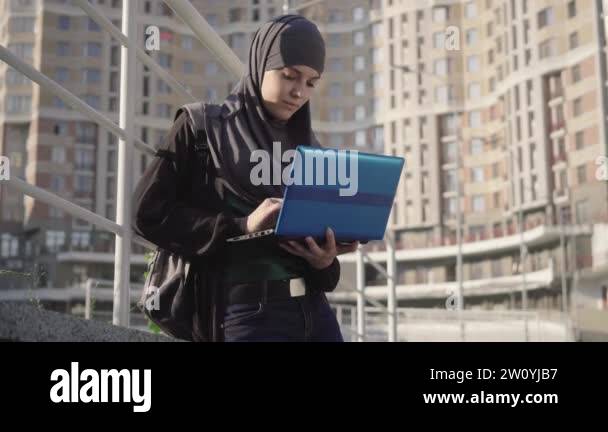 Smiling muslim woman in traditional hijab typing on laptop keyboard ...