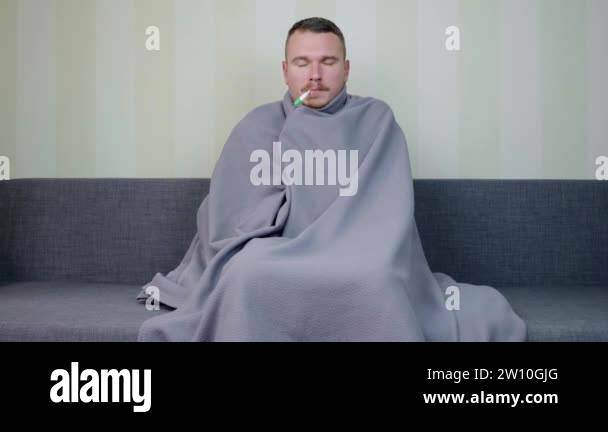 Sick man covered with blanket measures the temperature. Upset young guy ...
