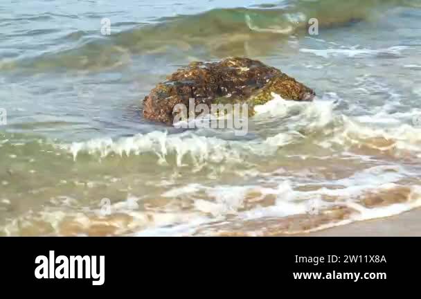 Sea waves flow around a small stone next to a sandy beach on a bright ...