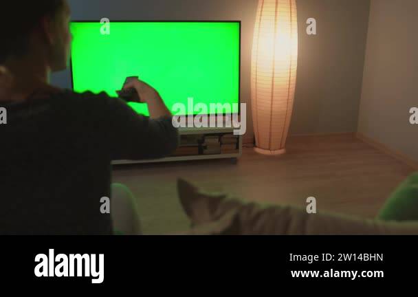 Woman is sitting on the couch, watching TV with a green screen, switching channels with a remote ...