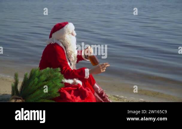 Bad Santa Claus sits with a bag of presents on the sandy shore of a ...