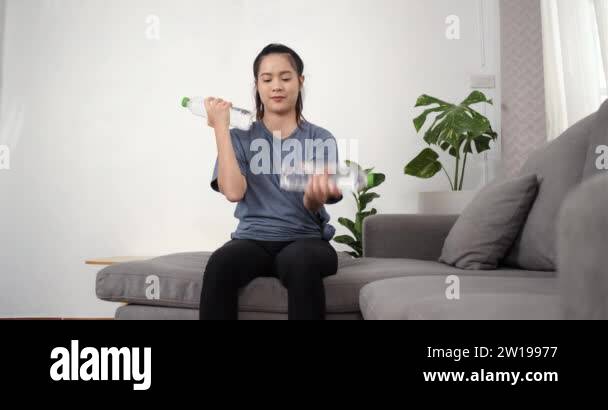 Home Workout of Young asian woman sitting down on sofa doing bicep curls with bottle of water as ...