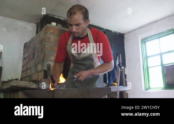 Cheery blacksmith is hitting a red bent billet with a hammer in a ...