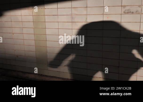 A large woman's shadow moves across a subway tile wall lit with sunlight as the shadow reveals a ...