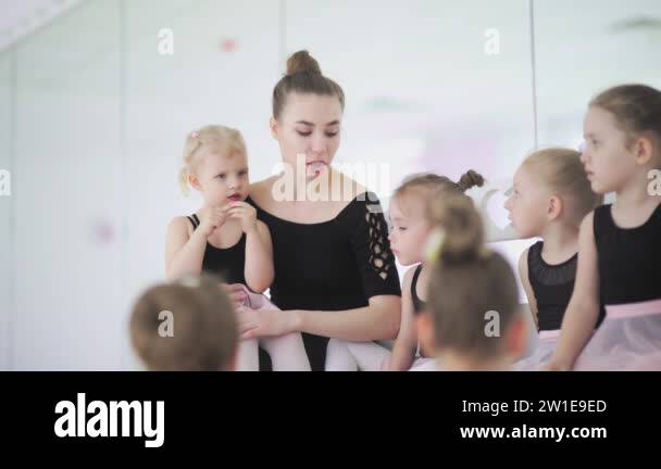 Young ballet teacher sitting on bench in ballet school with little ...