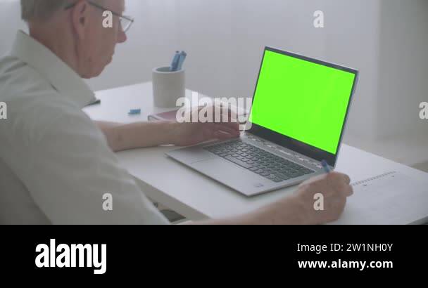 old man is looking at screen of laptop and writing notes in notebook ...