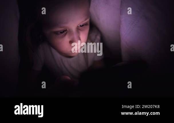 A young little girl is hiding under a blanket to use a digital tablet ...