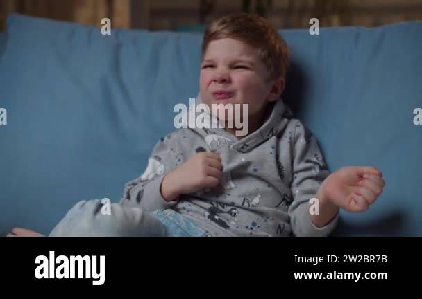 Blonde boy fooling around sitting on cozy sofa at evening room. Kid in ...