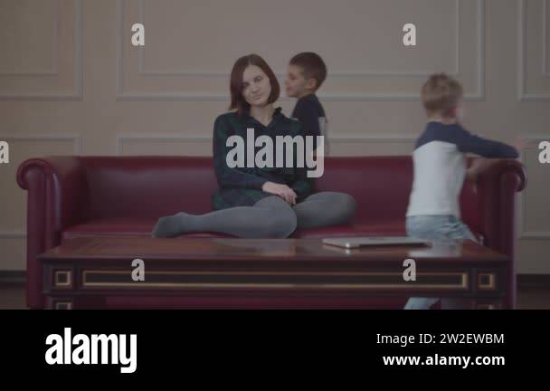 Exhausted young mother sitting on home red couch while two loud kids ...