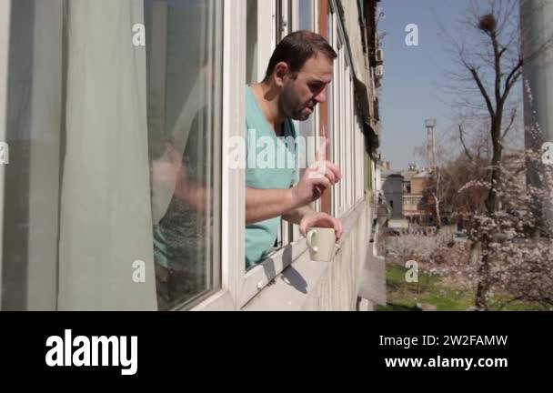 A disgruntled man stands near an open window with a cup in his hand ...