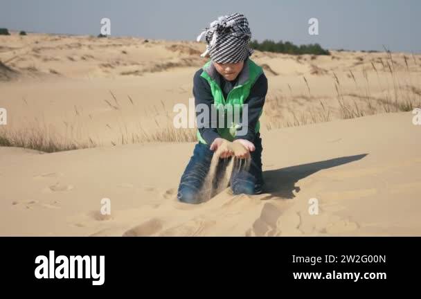 Cheery boy in shemagh, jeans and jacket playing with sand in desert in ...