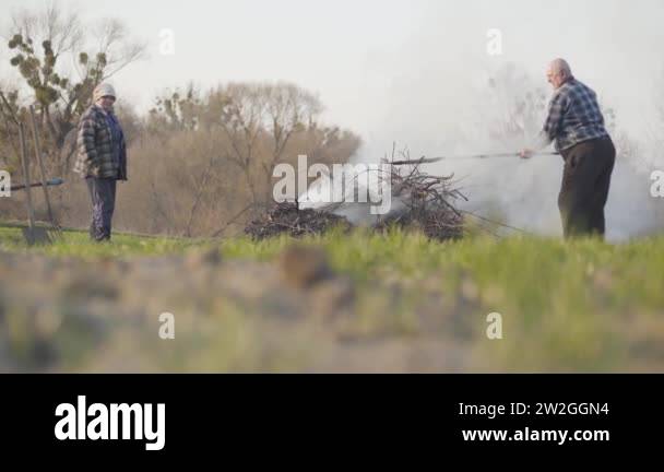 Wide shot of old Caucasian farmer using rake to mix the fire of burning ...