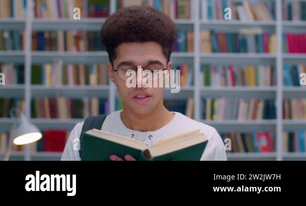 Close up of young Latino handsome guy in glasses standing and reading book in library. Good ...