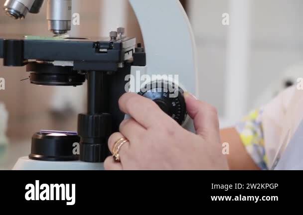 In close-up, the fingers slowly and gently rotate the microscope handle ...