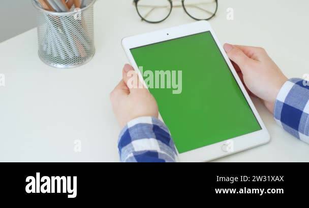 White vertical tablet device with green screen in hands of Caucasian woman at table. Female ...