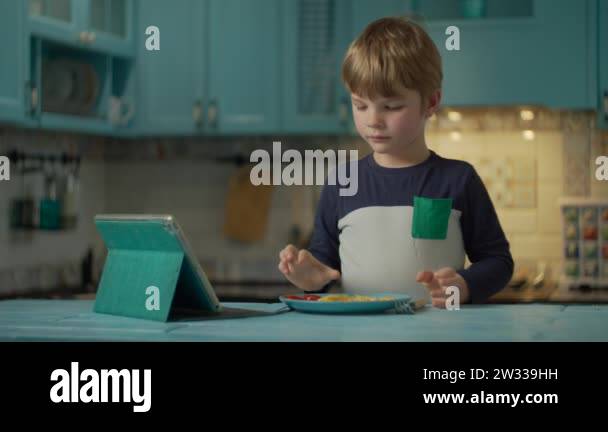 Young boy eating scrambled eggs with tomatoes watching tablet computer ...