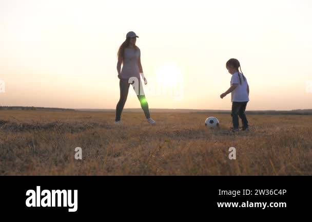 Mom and daughter play football on field in rays of the sunset. Teamwork ...