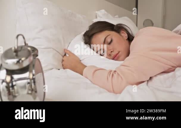 A young sleepy girl is turning off her alarm clock laying in the bed at ...