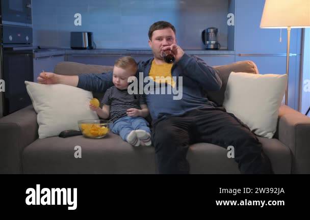 Family dad and son sit on couch and watch TV at home in evening and eat potato chips. Father ...