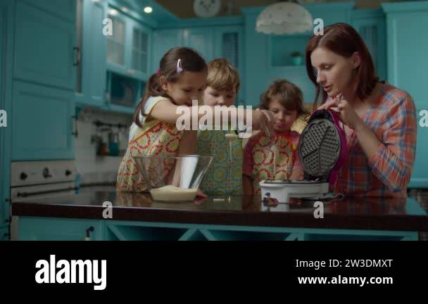 Young 30s mother with three kids in aprons making waffles in waffle ...