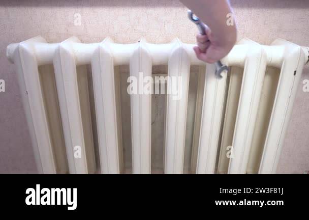 A man with a wrench knocks on the heating pipe of a radiator and does ...