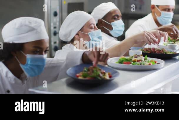 Diverse group of chefs wearing face masks garnishing dishes in ...
