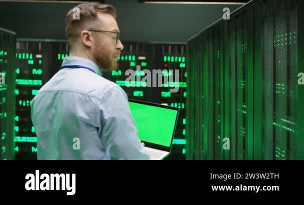 Rear of Caucasian young man in glasses walking among servers with laptop computer of green ...