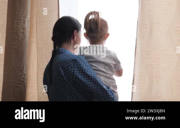 Mother stands with a small child looking out the window at home, the ...