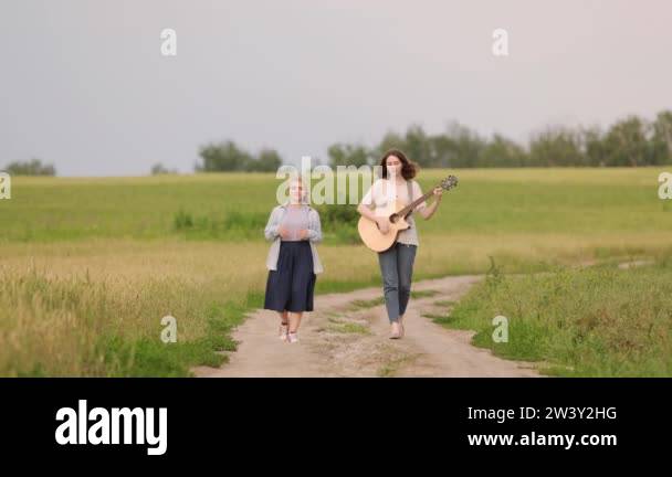 two young beautiful girls walk and sing along a wheat field at sunset ...