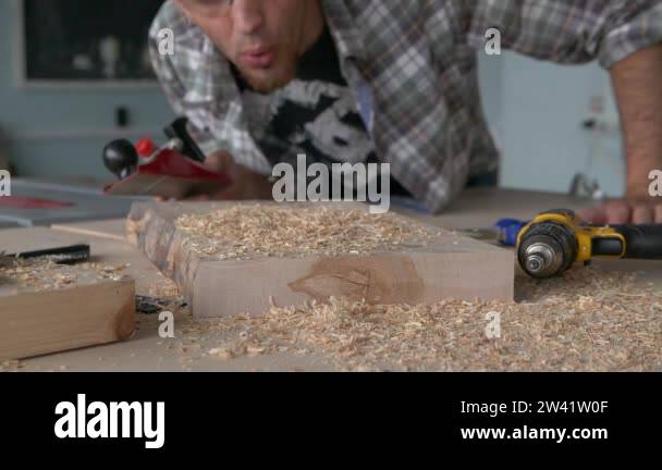 A young carpenter is cleaning a workbench from sawdust in a carpentry ...