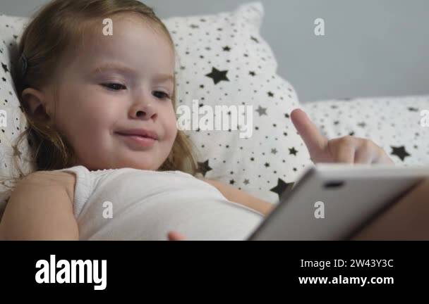 Little happy baby is playing tablet while lying on the bed. Kid watches ...